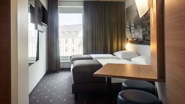 Quarto de hotel com cama, escrivaninha, cadeira, mesinha, janela com cortinas e TV fixada na parede.