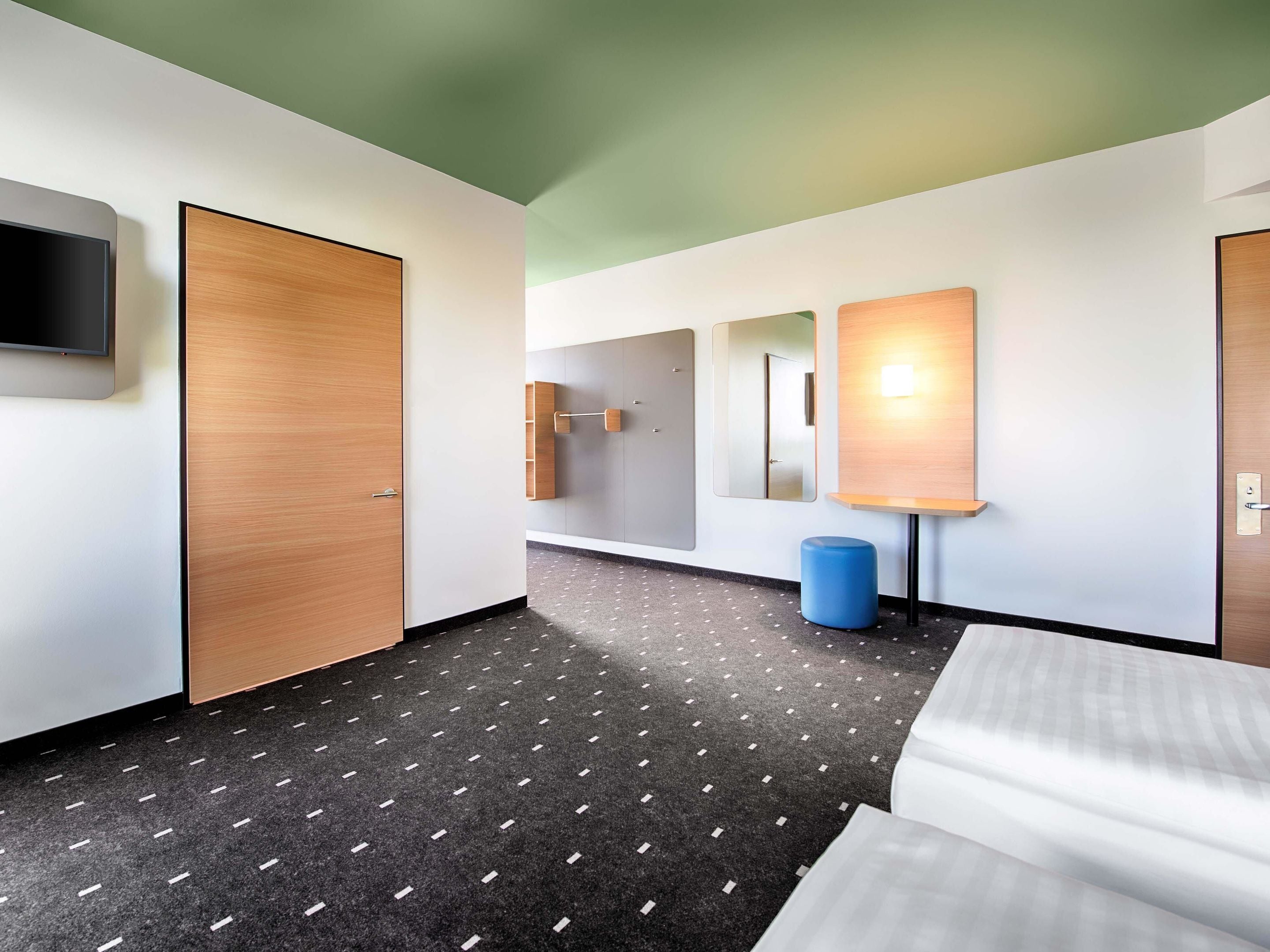 Quarto de hotel com uma cama, televisão, porta de madeira, espelho e uma lixeira azul.