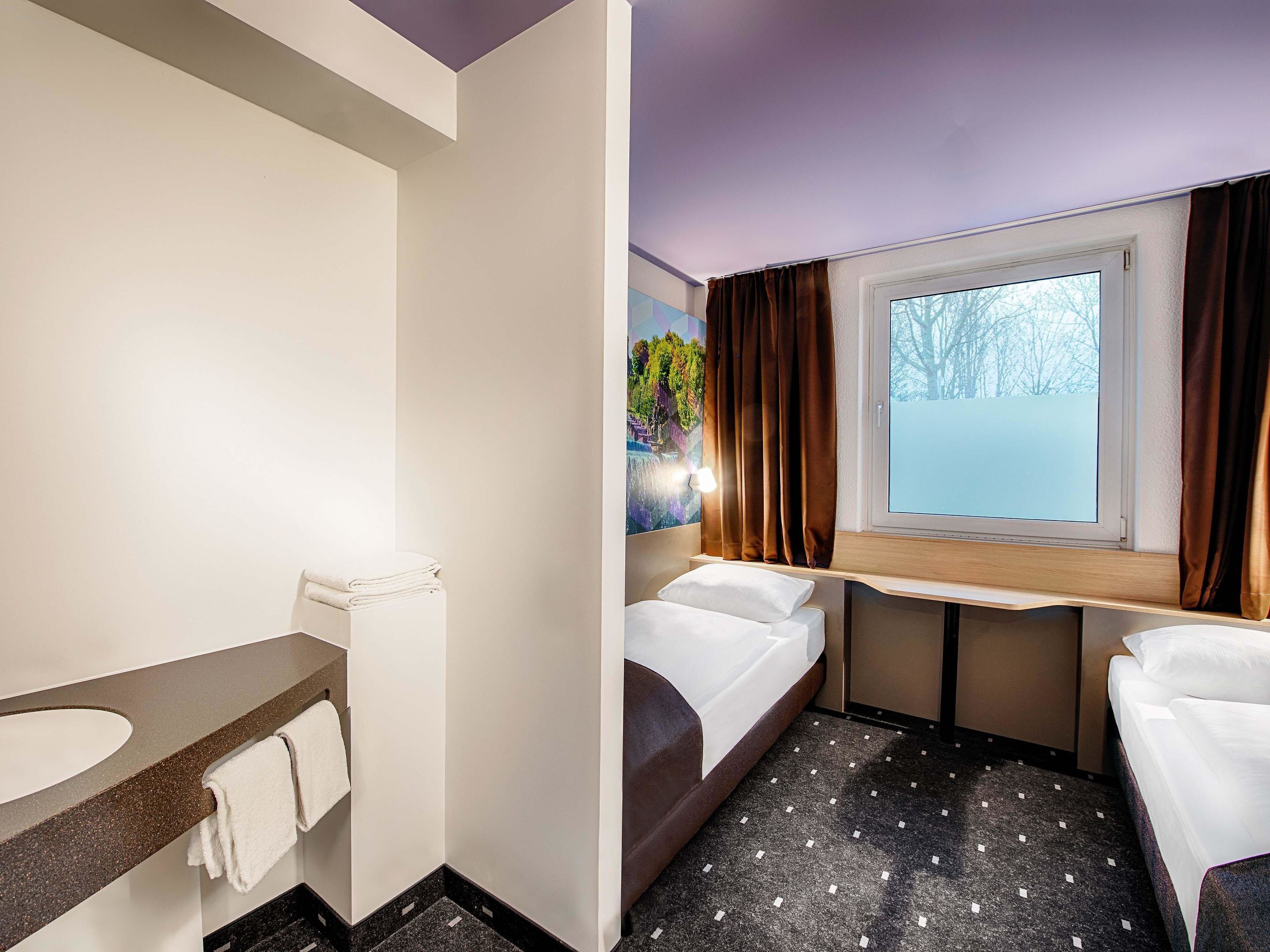 Quarto de hotel com cama, escrivaninha, janela com cortinas, um quadro na parede e banheiro com pia e toalha.