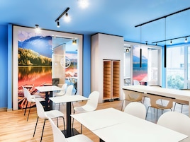 Uma área de refeições moderna com mesas e cadeiras brancas, uma janela grande com vista para um lago e uma parede com uma pintura panorâmica.