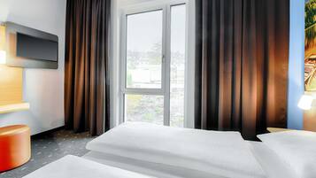 Quarto de hotel com cama, televisão fixada na parede, janela com cortinas e um quadro na parede.