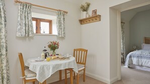 Uma área de refeições com uma mesa posta para dois, uma cadeira de madeira, uma janela com cortinas, uma estante com objetos decorativos e uma cama no quarto adjacente.
