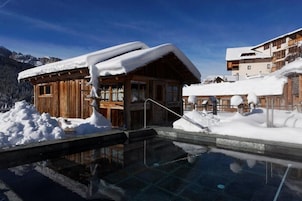 Um chalé de madeira com telhado coberto de neve, área de piscina e montanhas ao fundo.