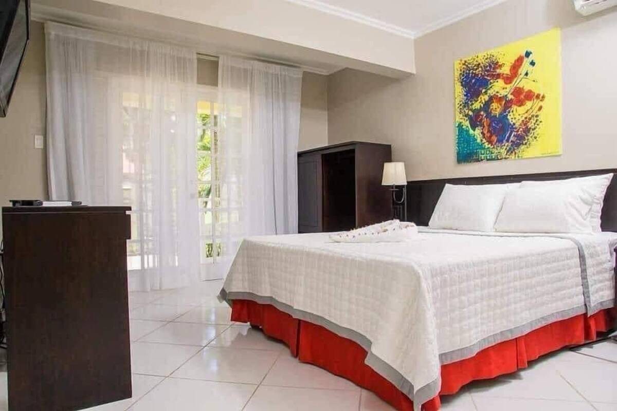 Quarto de hotel com cama, escrivaninha, luminária e uma pintura abstrata colorida na parede.