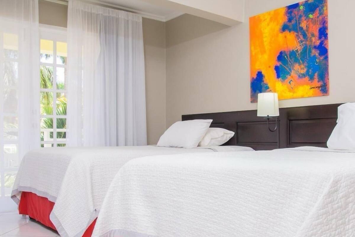 Quarto de hotel com duas camas, roupa de cama branca, base de cama vermelha e um grande quadro abstrato na parede, e uma janela com cortinas brancas.