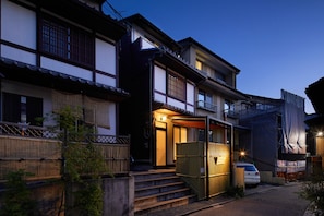Uma casa tradicional japonesa com escadas de madeira que levam à entrada, um carro estacionado na frente e um céu da noite claro.
