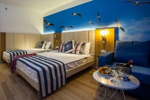 Quarto de hotel com duas camas, um sofá azul, uma mesinha com uma garrafa de champanhe e copos, e um mural de pássaros na parede.