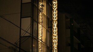 Fachada de edifício com sinalização vertical iluminada, padrão de janelas em grade e elementos estruturais visíveis.
