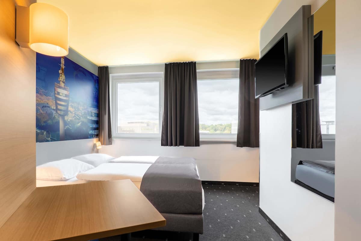 Quarto de hotel com cama, escrivaninha, televisão e janela com cortinas.