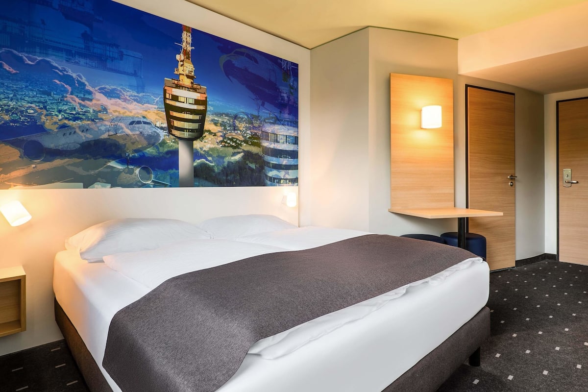 Quarto de hotel com uma cama grande, uma TV fixada na parede e uma porta com painel de madeira.