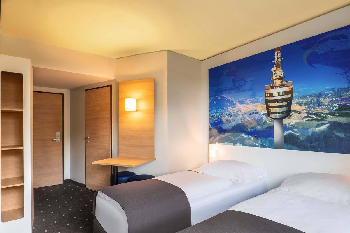 Quarto de hotel com duas camas, um guarda-roupa de madeira, uma secretária pequena e uma luminária na parede.