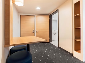 Quarto compacto com escrivaninha, duas cadeiras, guarda-roupa e banheiro visível através de uma porta de vidro.