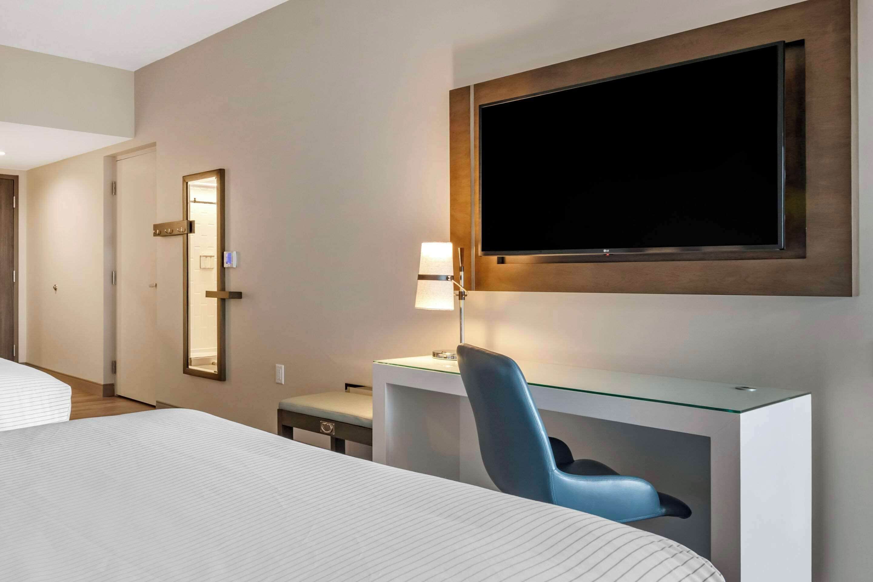 Quarto de hotel com uma cama grande, uma escrivaninha com cadeira, uma televisão na parede e um espelho sobre a escrivaninha.