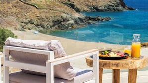 Vista à beira-mar com uma cadeira branca e uma mesa com comida e bebidas, com vista para o mar azul cristalino e colinas rochosas.