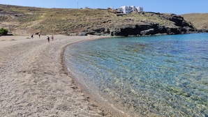 Uma praia com águas cristalinas e rasas, uma costa rochosa e um edifício numa colina ao fundo.