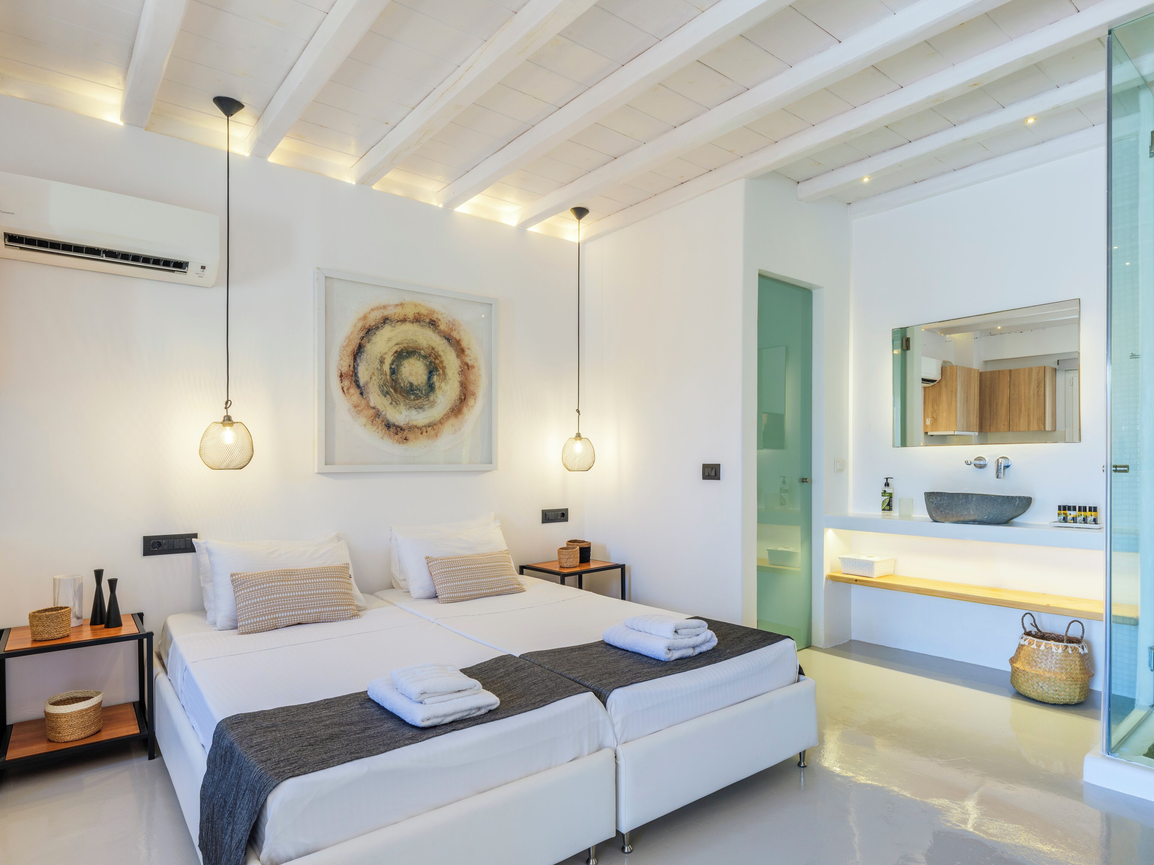 Quarto moderno com cama de casal, mesas de cabeceira, uma grande pintura abstrata, banheiro com pia e espelho, e box de vidro.
