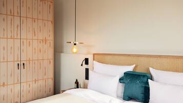 Quarto de hotel com um guarda-roupa espaçoso, cama com roupa de cama branca, cabeceira de madeira e uma lâmpada suspensa.