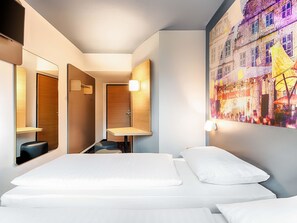 Quarto de hotel com cama, espelho, pequena escrivaninha e televisão na parede.