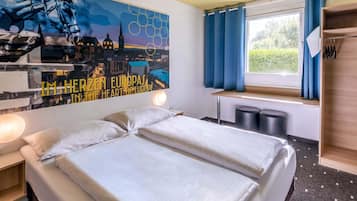 Quarto de hotel com cama, mesa de cabeceira, arte na parede retratando uma paisagem urbana, janela com cortinas e prateleira de madeira com objetos.