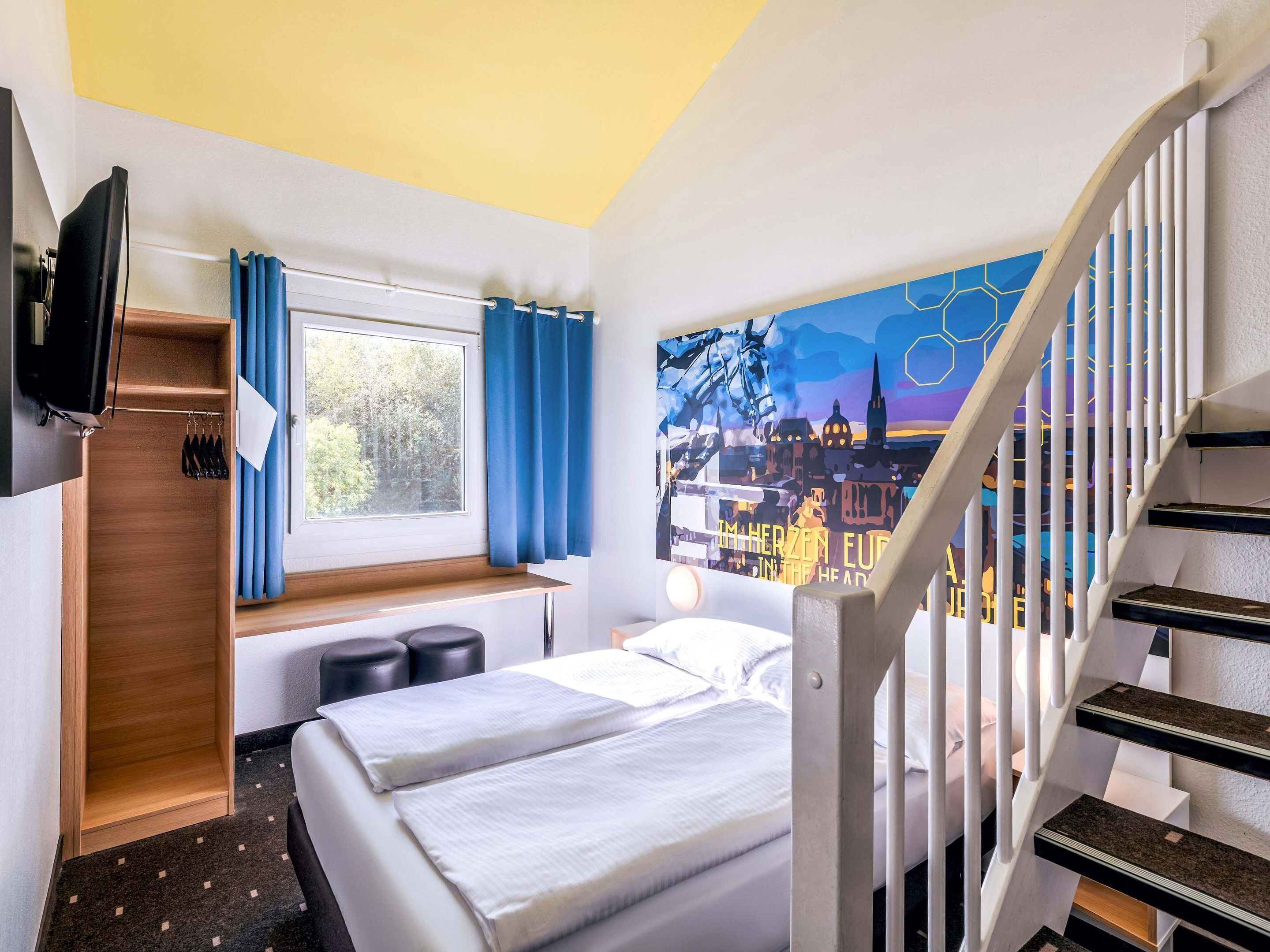 Quarto de hotel com cama, escrivaninha, janela com cortinas, um quadro na parede e uma escada.