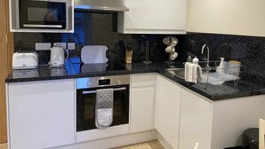 Cozinha compacta com armários brancos, bancada preta, micro-ondas, torradeira, chaleira e pia com porta-toalhas.