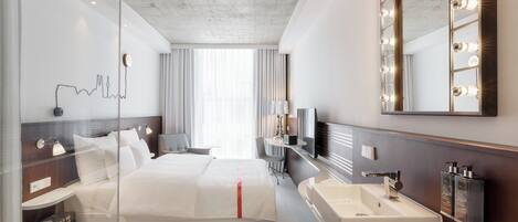 Quarto de hotel com cama, mesa de cabeceira, luminária, espelho iluminado e banheiro com pia e espelho.
