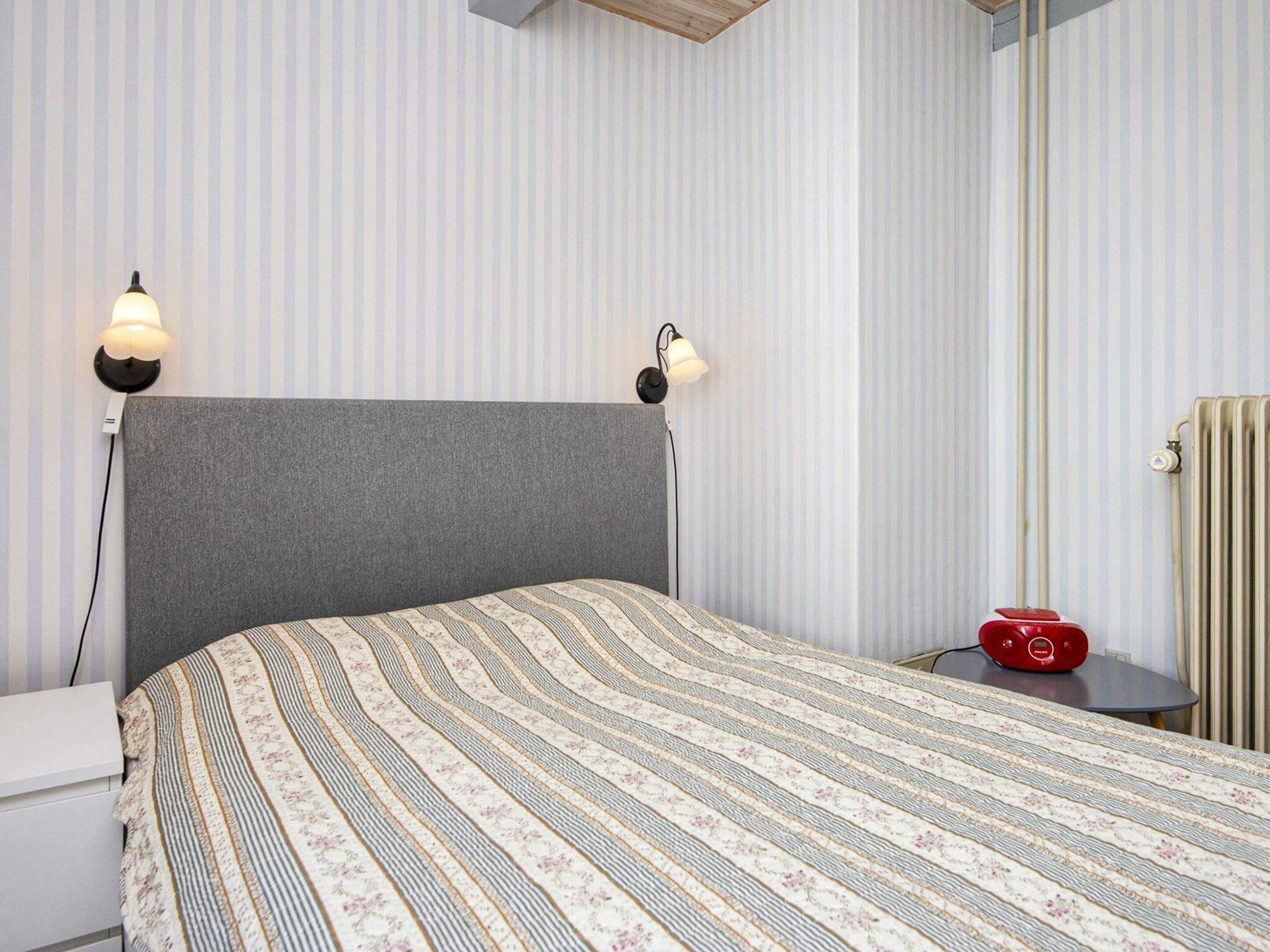 Quarto com cama, duas luminárias laterais, radiador e um despertador vermelho sobre uma mesinha de cabeceira.