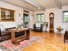 Um quarto com piso de madeira, um sofá de couro marrom, uma mesa de centro de madeira, uma grande pintura de paisagem, um relógio de chão e uma janela com vista para o verde.