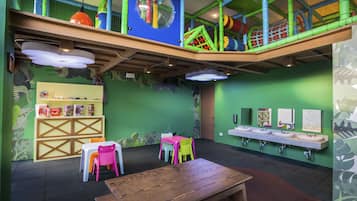 Uma área de recreação infantil com uma estrutura de escalada, uma mesa com cadeiras e uma estante de livros infantis.