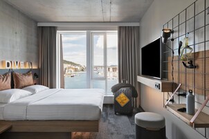 Quarto de hotel com uma cama, televisão, mala e vista para a cidade através de grandes janelas.