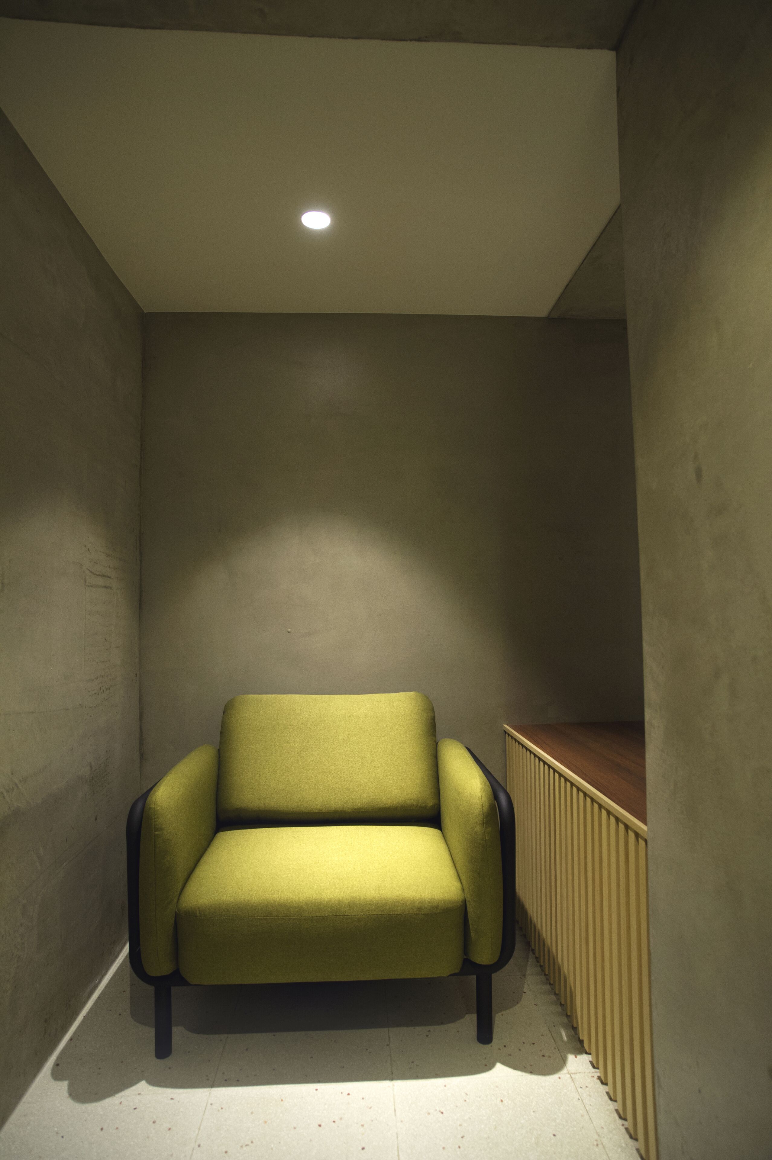 Um quarto pequeno e com pouca luz, com uma poltrona verde solitária, um radiador e paredes simples.