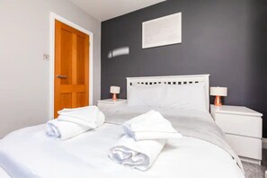 Cama bem arrumada com lençóis e toalhas brancos, mesa de cabeceira branca com abajur e porta de madeira.
