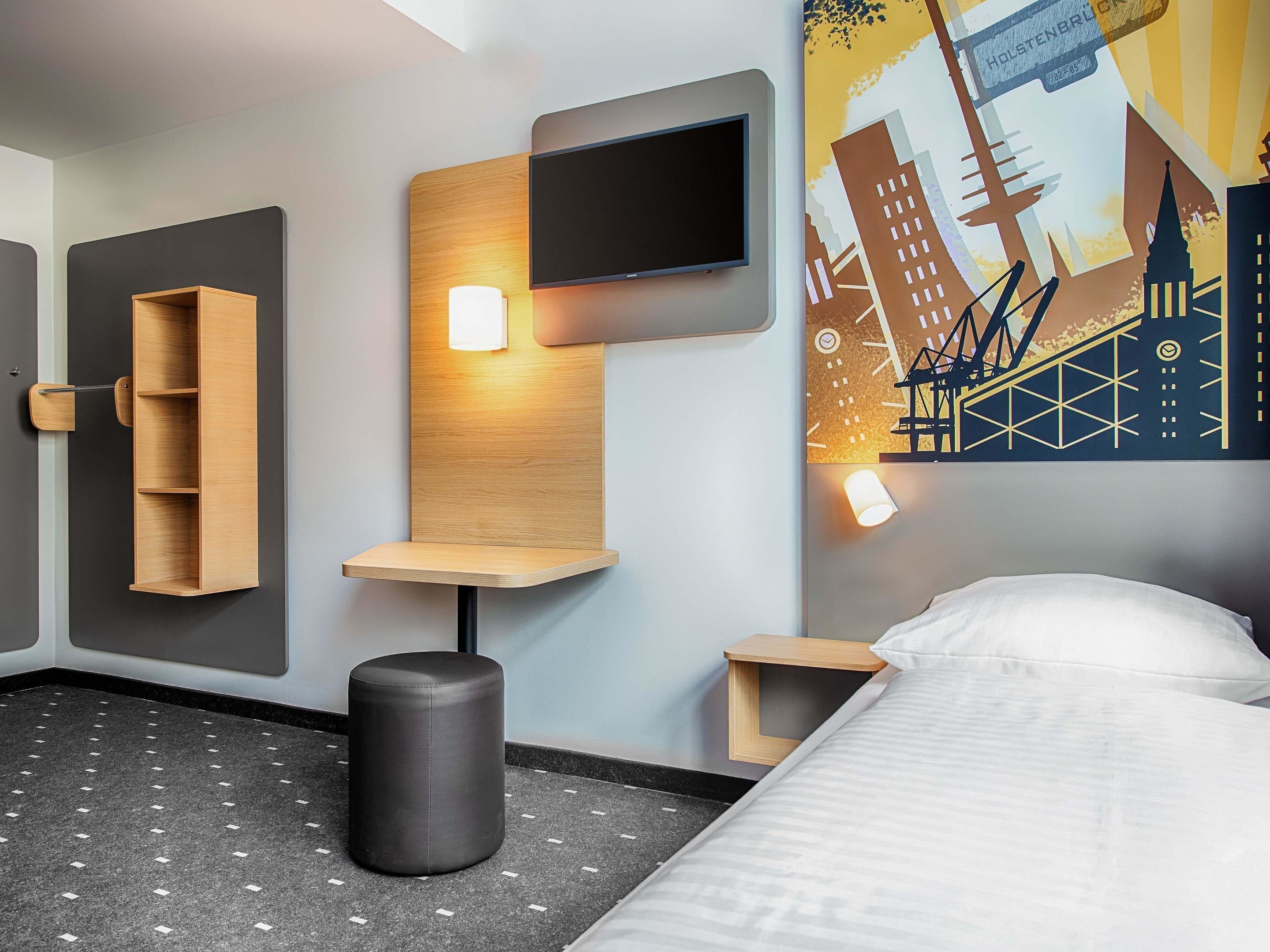 Quarto de hotel moderno com cama, televisão montada na parede, estante de madeira, um banquinho e luminária fixada na parede.