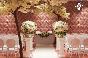 Decoração de cerimônia de casamento com cadeiras brancas, arranjo floral no altar e parede com padrão geométrico.