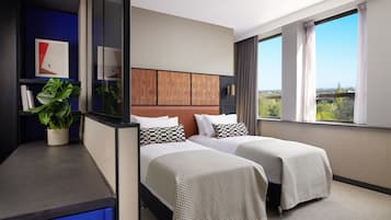 Quarto de hotel com duas camas, uma janela grande com vista para a cidade, uma planta sobre uma mesinha de cabeceira e uma parede azul decorativa.