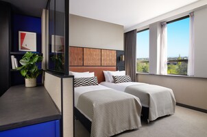 Quarto de hotel com duas camas, uma janela grande com vista para a cidade, uma planta sobre uma mesinha de cabeceira e uma parede azul decorativa.