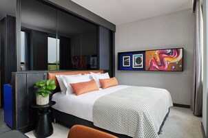 Quarto de hotel moderno com uma cama grande, uma pequena mesa lateral com uma planta, um espelho amplo e arte abstrata na parede.