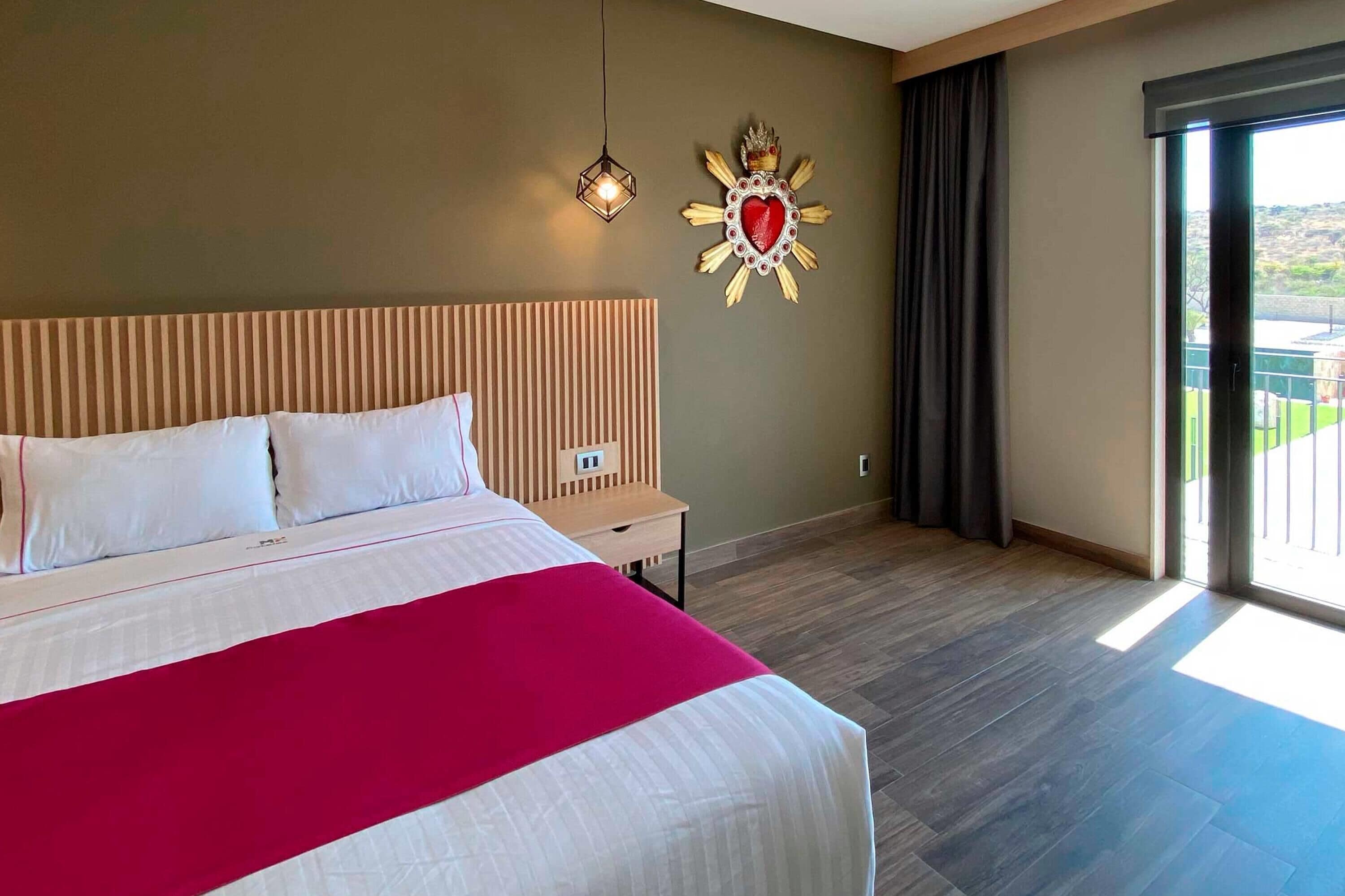Quarto de hotel com uma cama grande, mesa de cabeceira, luminária fixada na parede e um objeto decorativo em formato de coração.