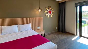 Quarto de hotel com uma cama grande, mesa de cabeceira, luminária fixada na parede e um objeto decorativo em formato de coração.