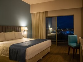 Quarto de hotel com uma cama grande, uma mesa com cadeira, uma janela com vista para a cidade e uma cortina.