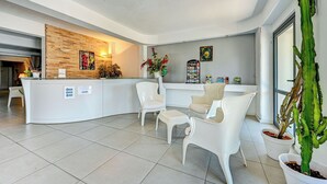 Uma área de recepção com balcão branco, várias cadeiras brancas e uma grande planta de cacto.