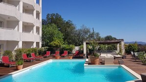 Uma área de piscina com espreguiçadeiras, uma pérgula e um edifício com varandas.