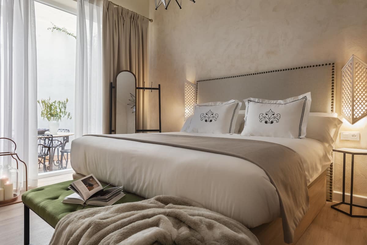 Quarto bem arrumado com uma cama grande, mesa de cabeceira com luminária, janela com cortinas e vista para o exterior.