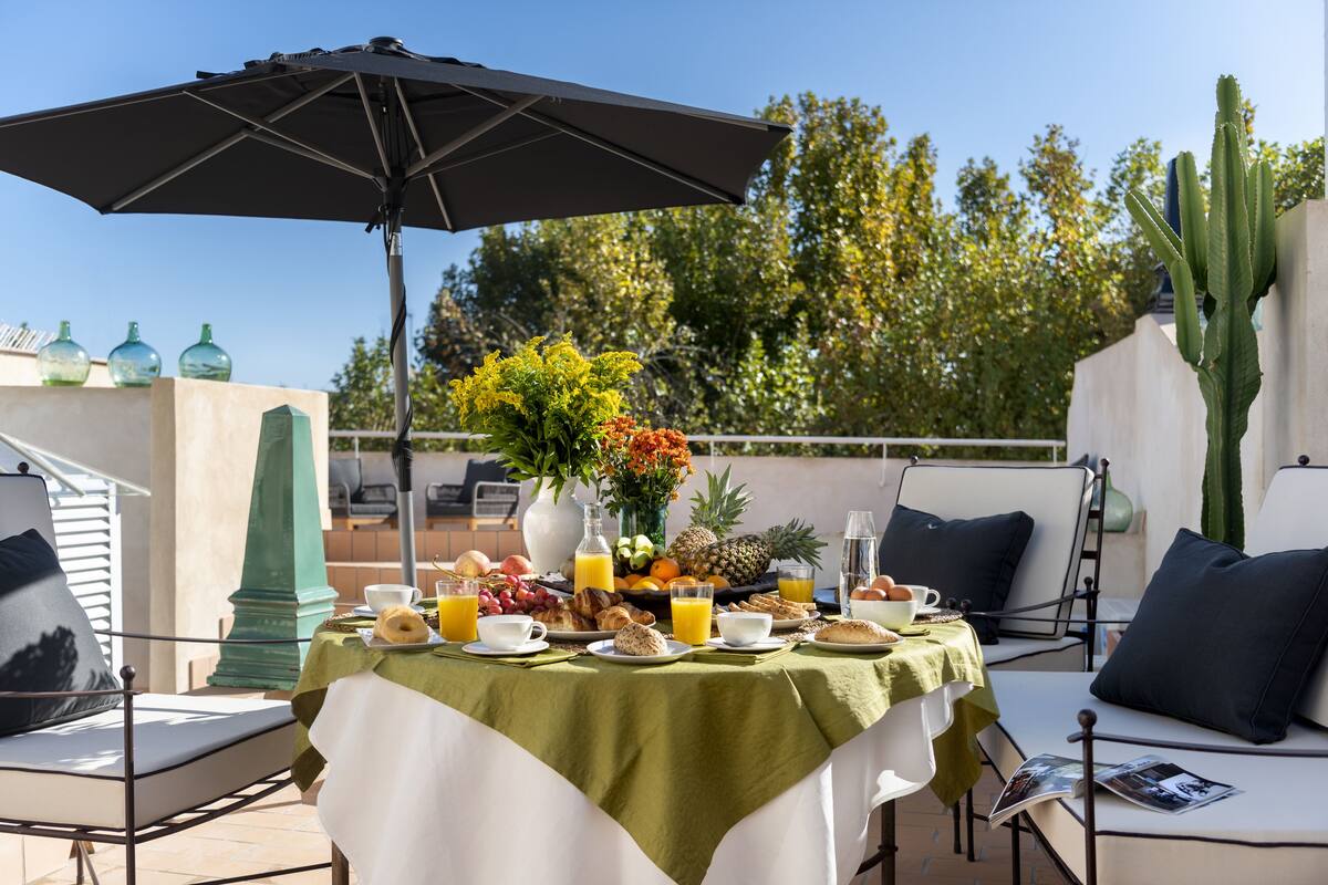 Mesa posta para o pequeno-almoço com toalha de mesa verde, cadeiras brancas e um guarda-sol preto.
