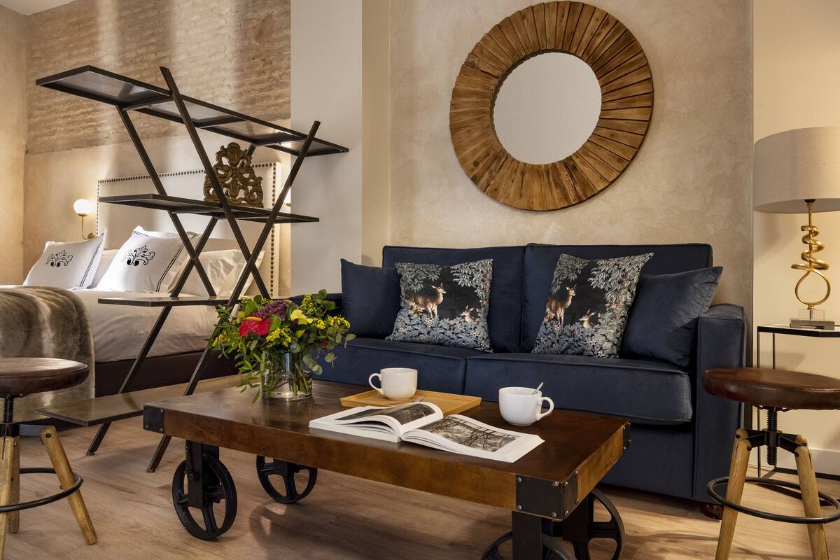 Uma sala de estar moderna com um sofá azul, uma mesa de centro de madeira com um livro e uma xícara, e um espelho decorativo na parede.