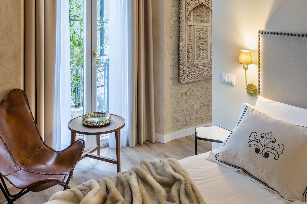 Quarto de hotel com cama, uma cadeira, uma mesinha, uma luminária e vista para o verde lá fora.
