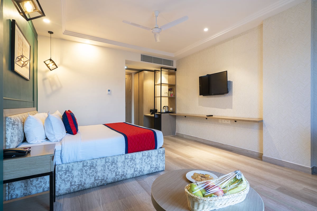 Quarto de hotel com cama, mesa de cabeceira, luminária, TV de tela plana e uma pequena mesa de jantar com uma cesta de frutas.