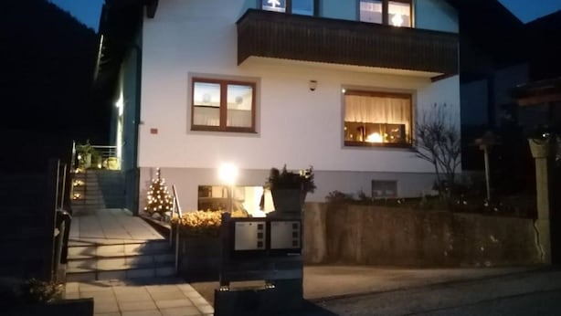 Uma casa de dois andares com varanda, uma escadaria que leva à entrada e um pequeno pinheiro de Natal no jardim frontal.
