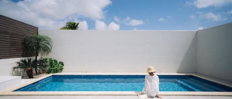 Piscina com uma pessoa sentada na beira, uma espreguiçadeira e uma pequena mesa com uma garrafa.
