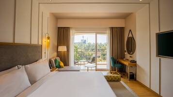 Quarto de hotel com uma cama grande, uma escrivaninha com cadeira, televisão e varanda com vista para áreas verdes e edifícios.